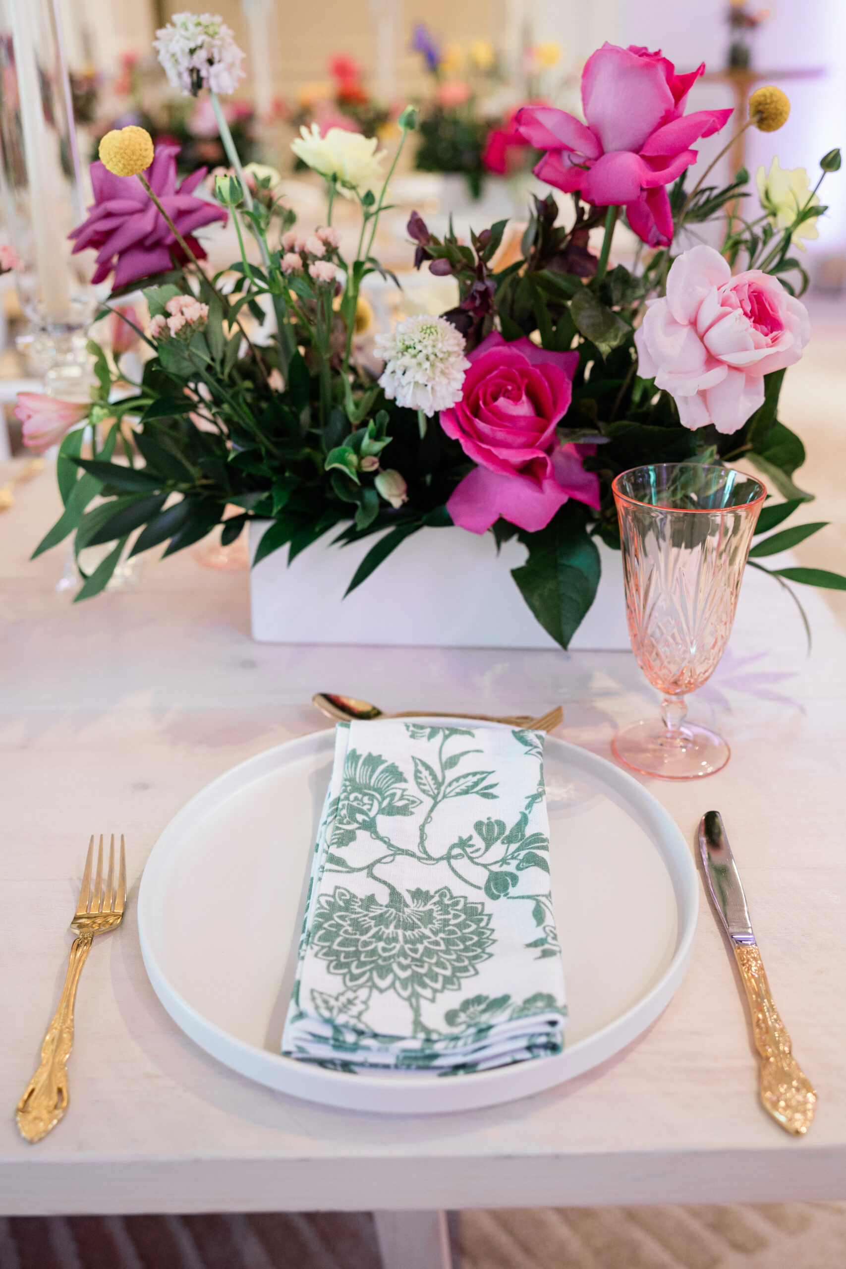 A white plate with a green floral napkin, gold utensils, and a pink ...