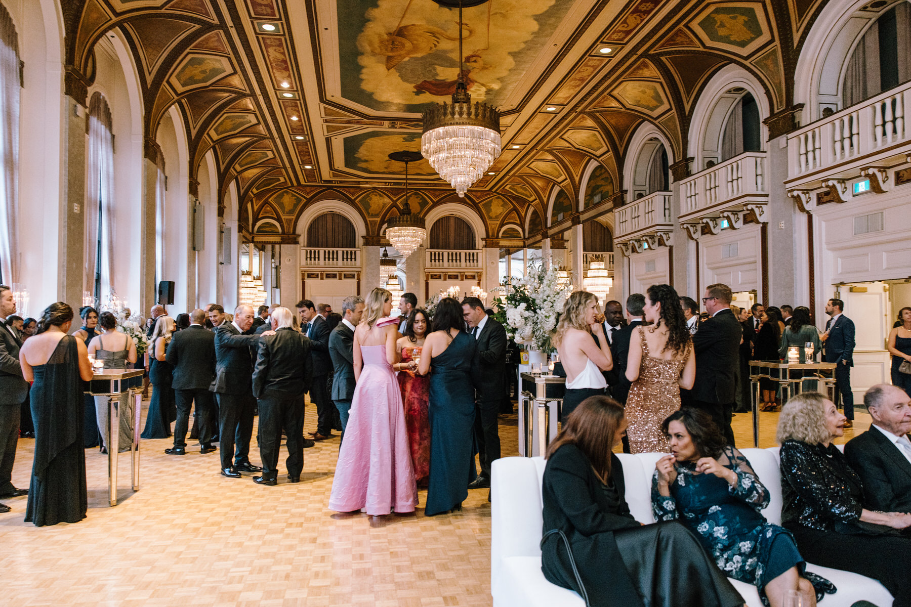A large group of elegantly dressed people mingle and chat in a grand ...