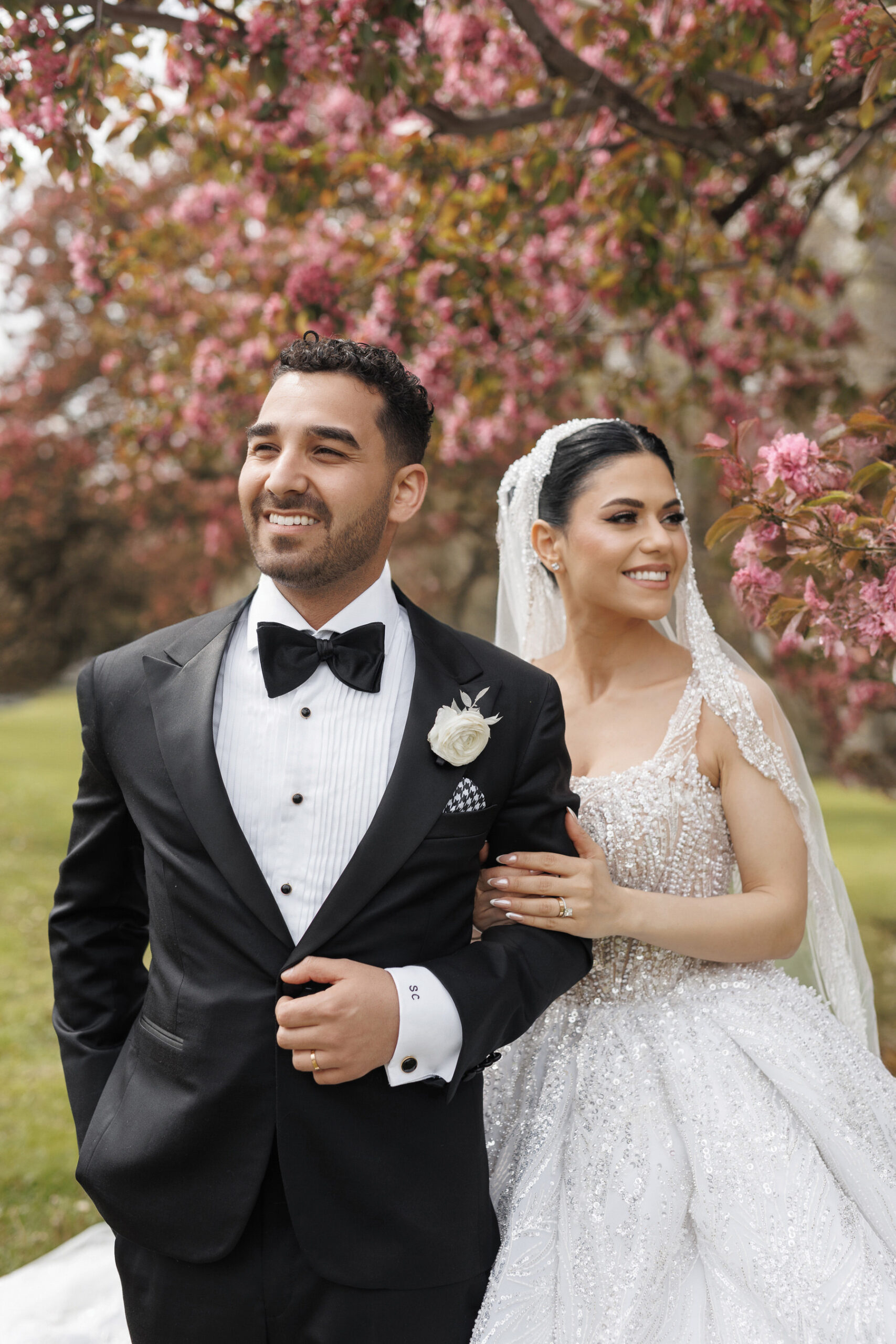 A smiling bride and groom stand outdoors under blooming pink trees ...