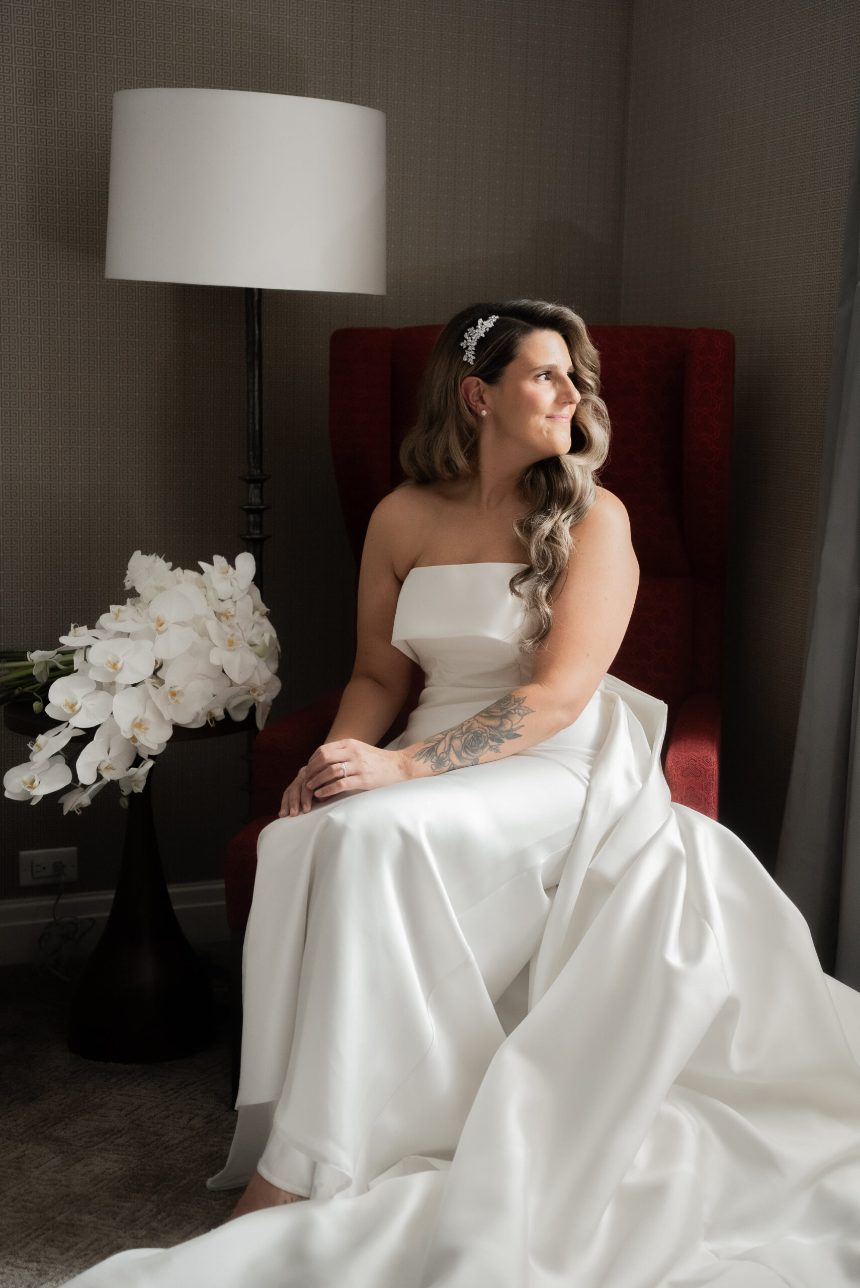 A bride in a strapless white gown sits on a red chair, smiling to the ...