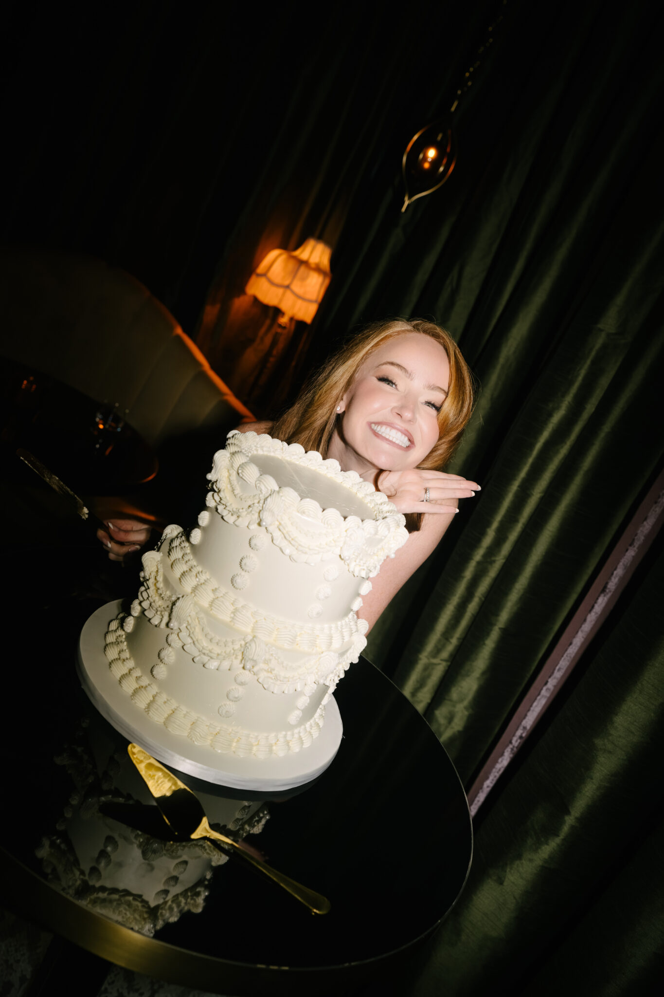 A smiling woman poses playfully behind a tall, ornate white cake with ...