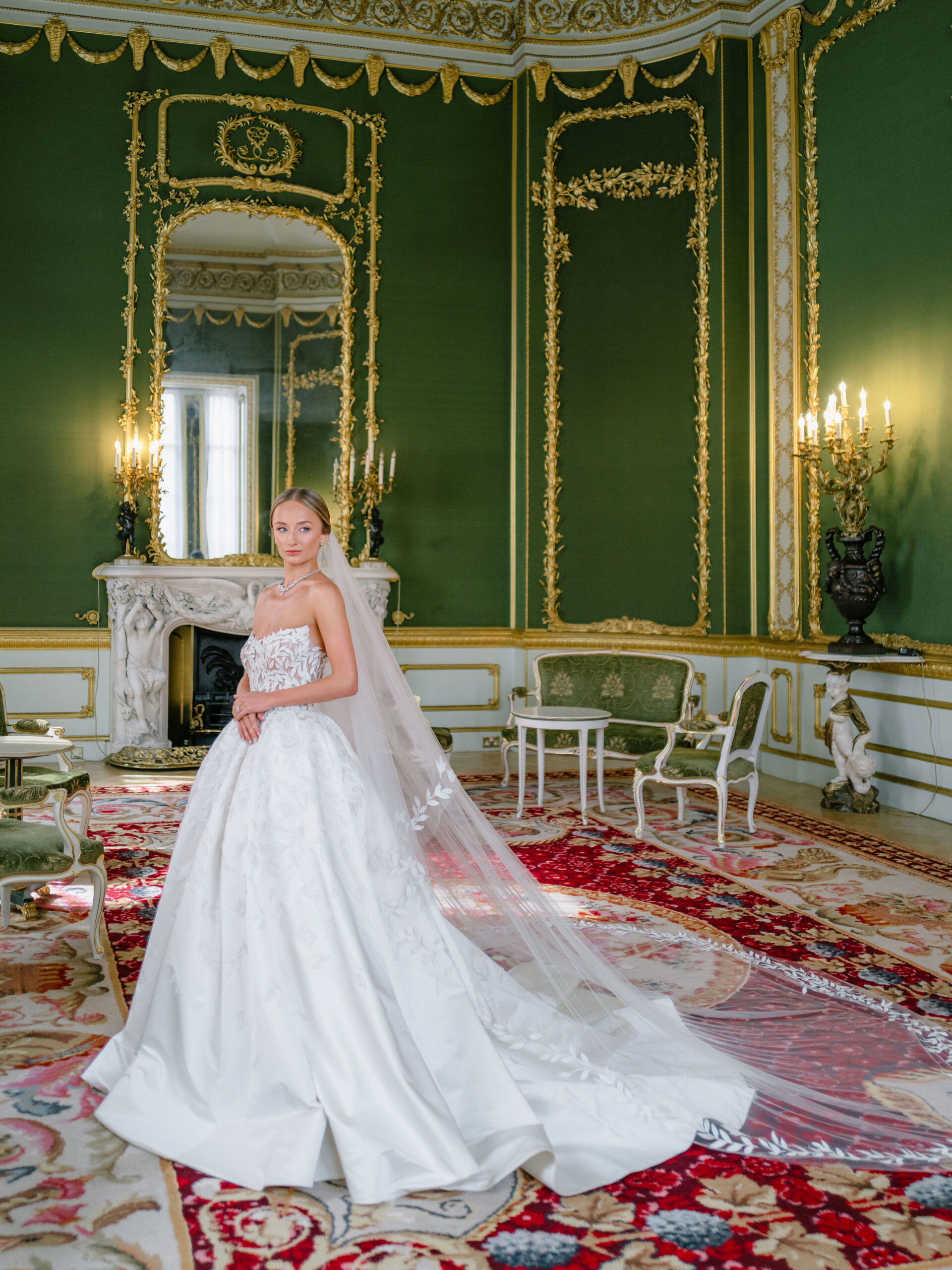 A bride in a strapless white gown with a long train poses in an ornate ...