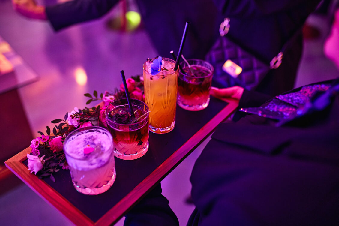 A person holds a tray with four colorful cocktails garnished with ...
