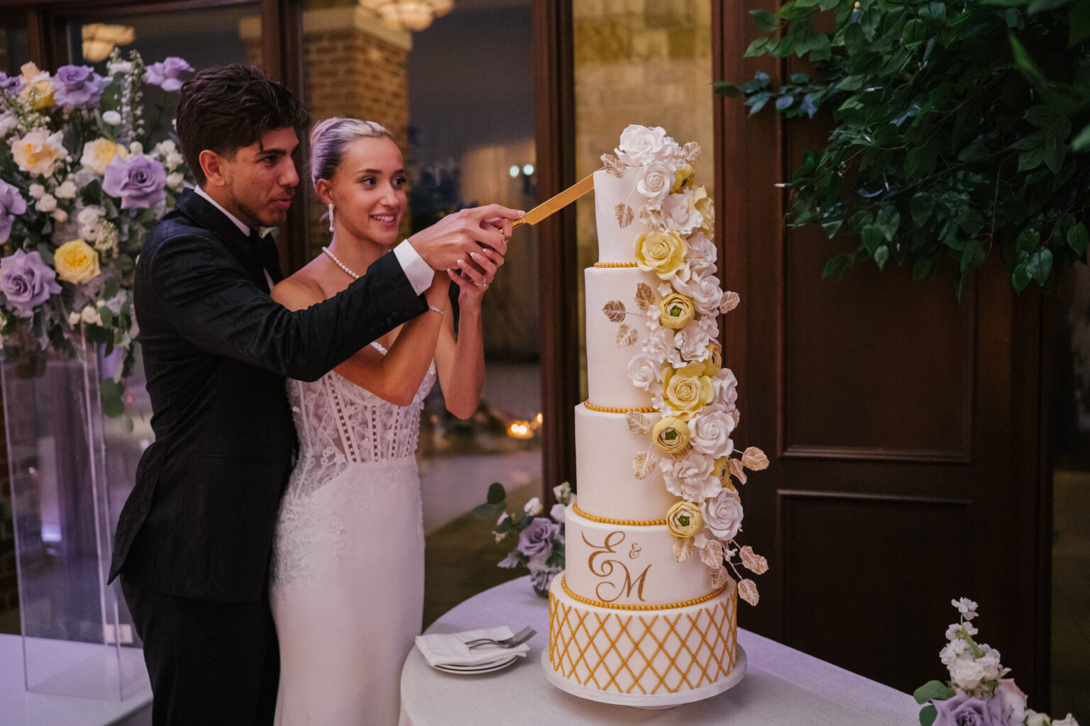A bride and groom smile as they cut a tall, elegant white wedding cake ...