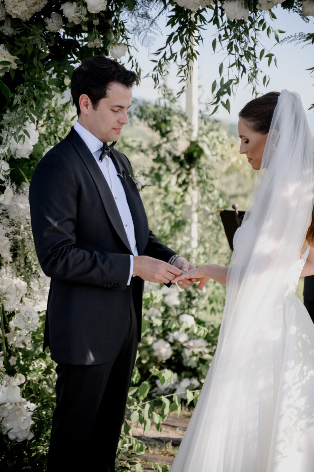 A groom in a black tuxedo places a ring on the bride’s finger during an ...