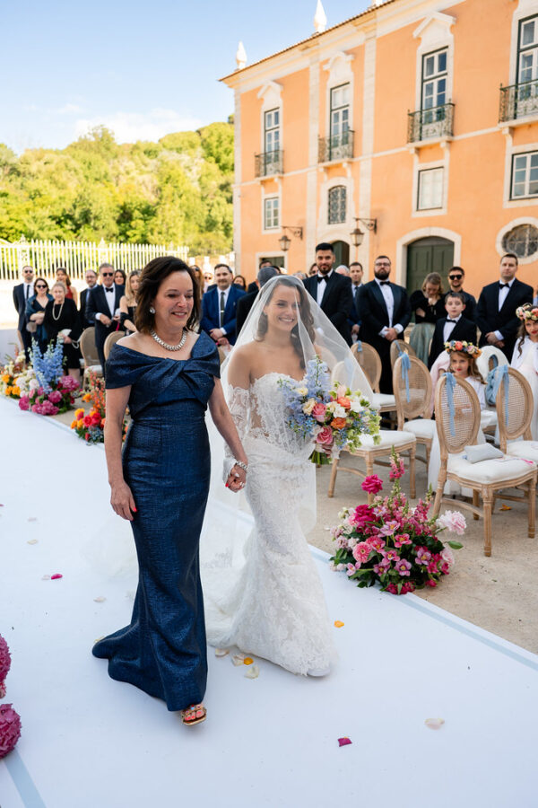 A bride in a white lace gown and veil walks down the aisle holding ...