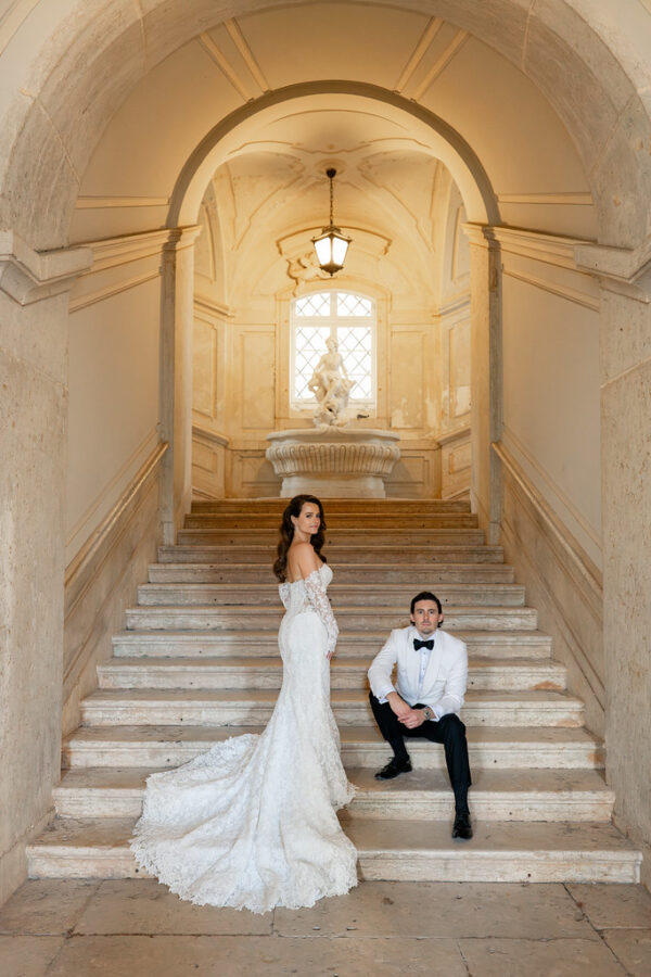 A bride in a white lace gown stands on stairs beside a groom in a white ...